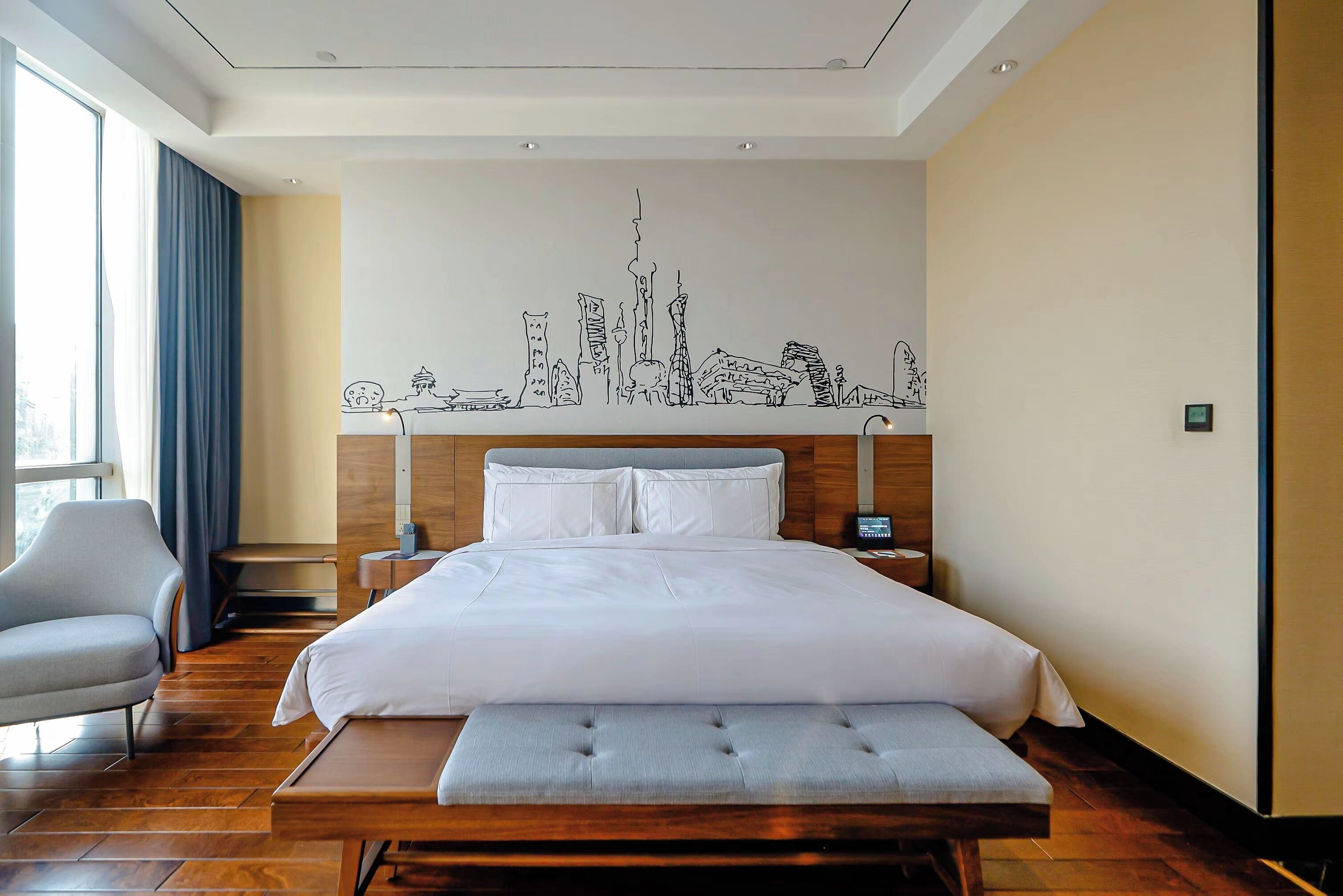 Quarto de hotel com uma cama grande, uma mesa de cabeceira, uma cadeira, piso de madeira e arte na parede com vista para a cidade.