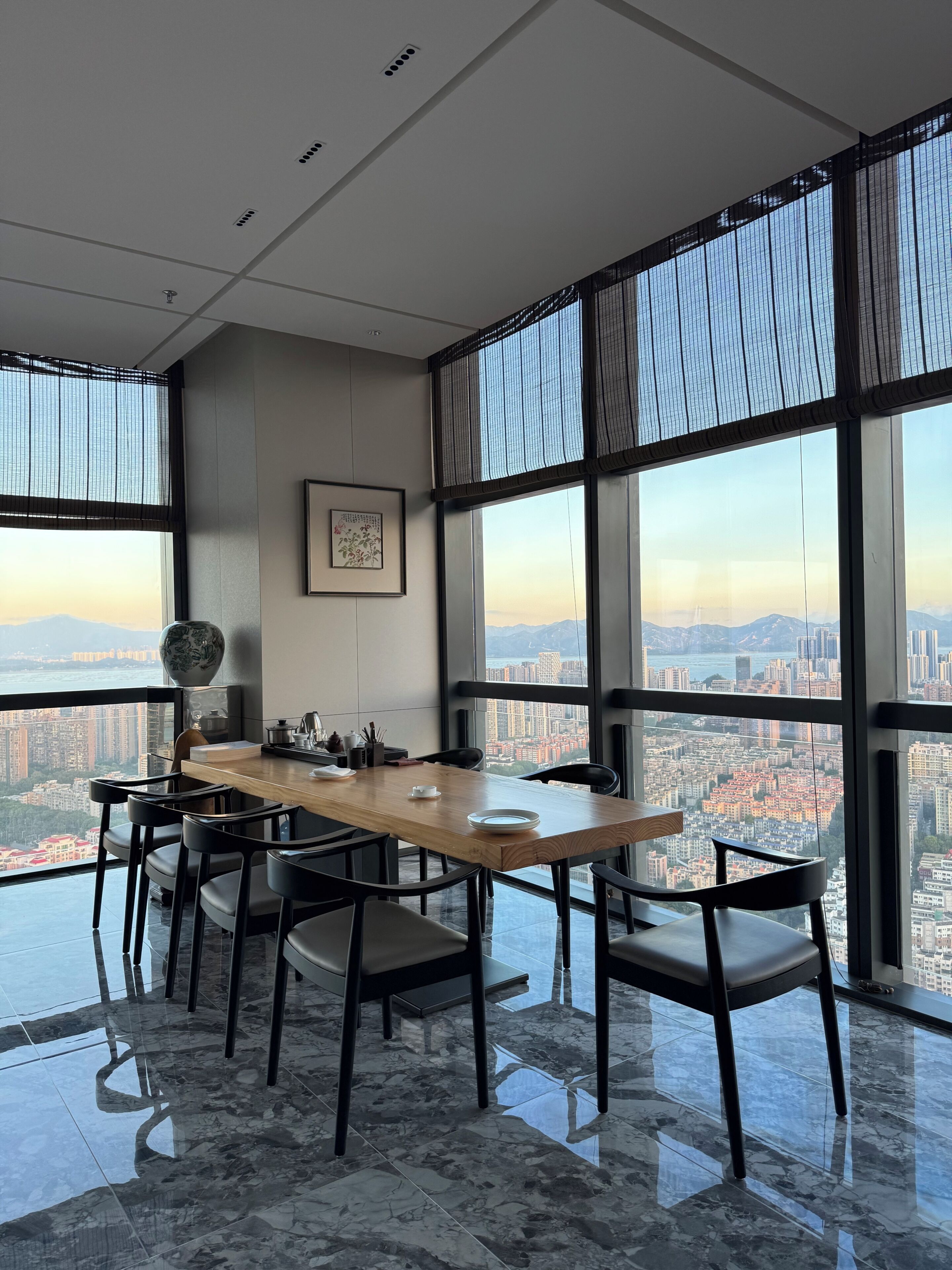 Uma área de refeições em um prédio alto com vista panorâmica da cidade, uma mesa de madeira e cadeiras modernas.