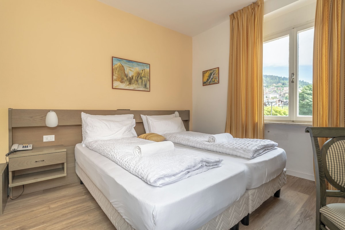 Quarto de hotel com duas camas, uma mesa de cabeceira com abajur, uma cadeira e vista para a cidade pela janela.