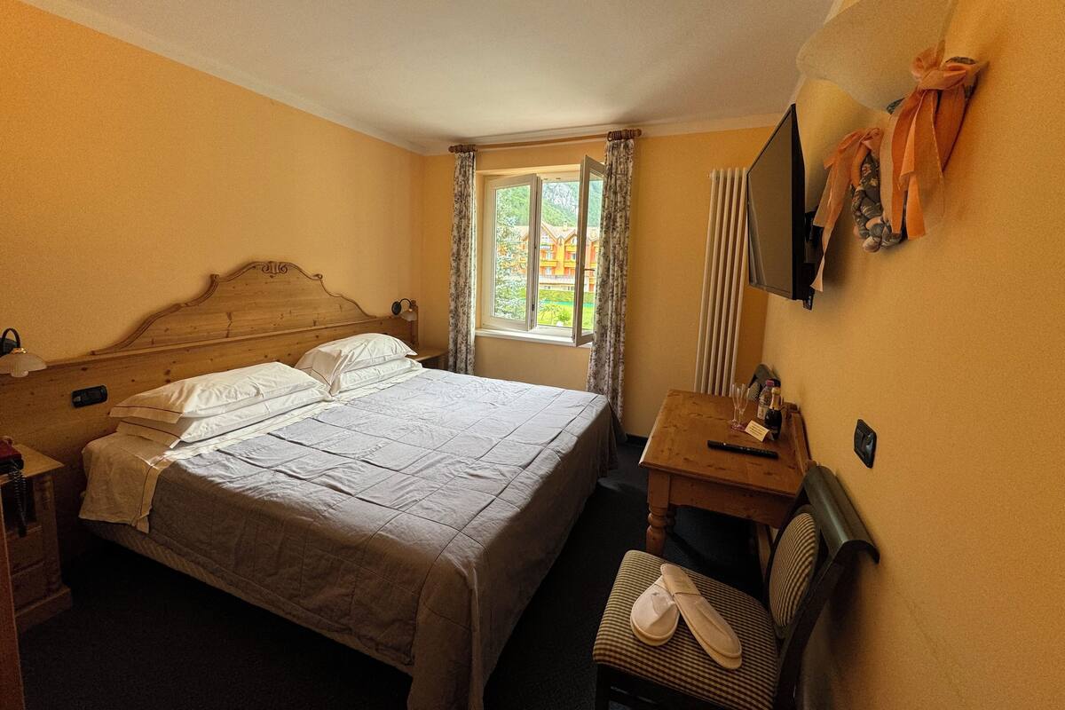 Quarto de hotel com cama, escrivaninha, cadeira, janela com cortinas e televisão na parede.