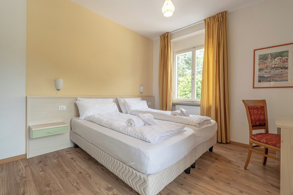 Quarto de hotel com cama, uma cadeira, janela com cortinas, luminária na parede e um quadro.