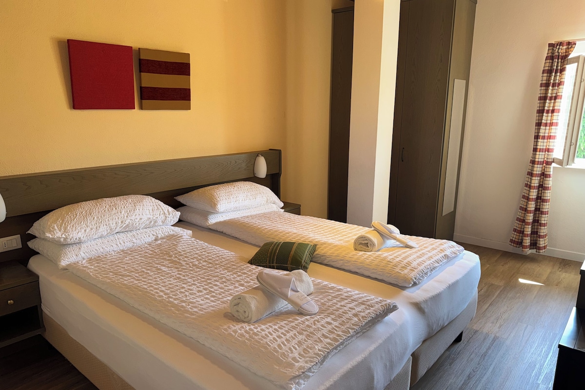 Quarto de hotel com duas camas, cabeceira de madeira, luminária fixada na parede e janela com cortinas.