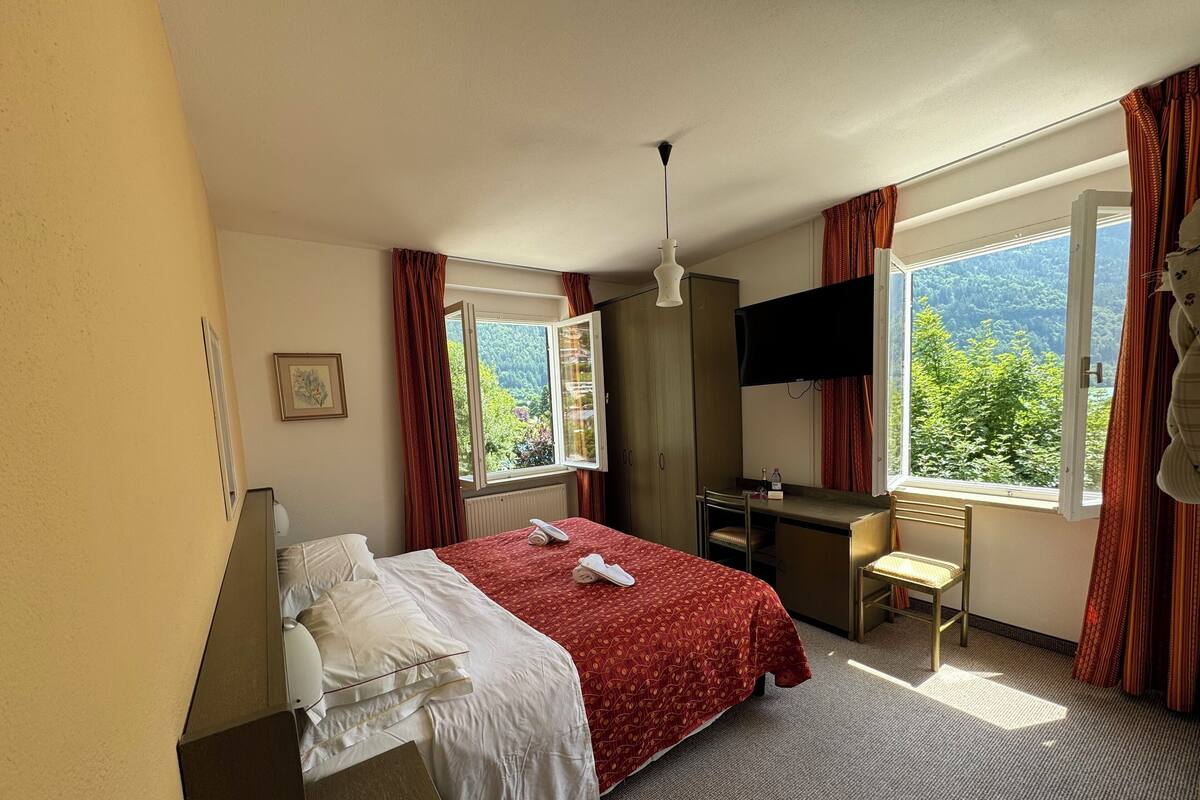 Quarto de hotel com cama, escrivaninha e grandes janelas com vista para áreas verdes.