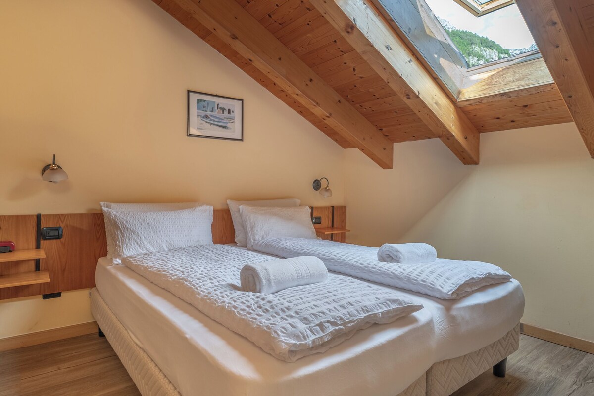 Quarto com teto de madeira, cama com roupa de cama branca, mesa de cabeceira e abajur fixado na parede.