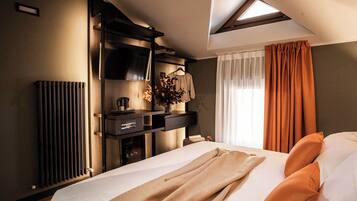 Quarto de hotel com cama, escrivaninha, radiador, janela com cortinas e claraboia.