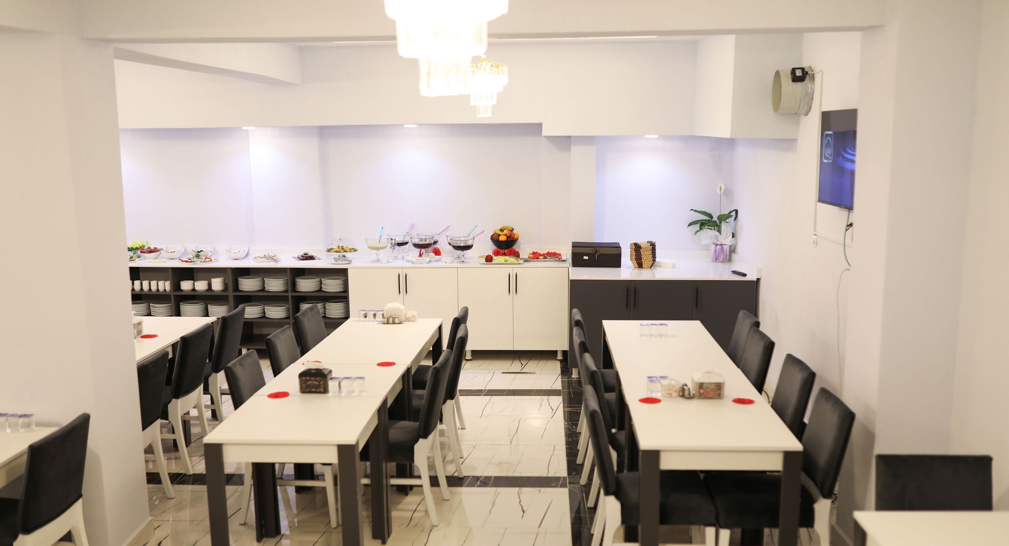 Uma sala de jantar moderna com mesas brancas e cadeiras pretas, um candelabro e uma cozinha ao fundo.