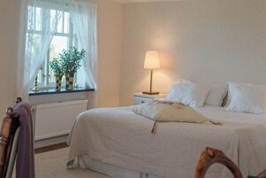 Quarto com cama, mesa de cabeceira, luminária, janela com cortinas e radiador.