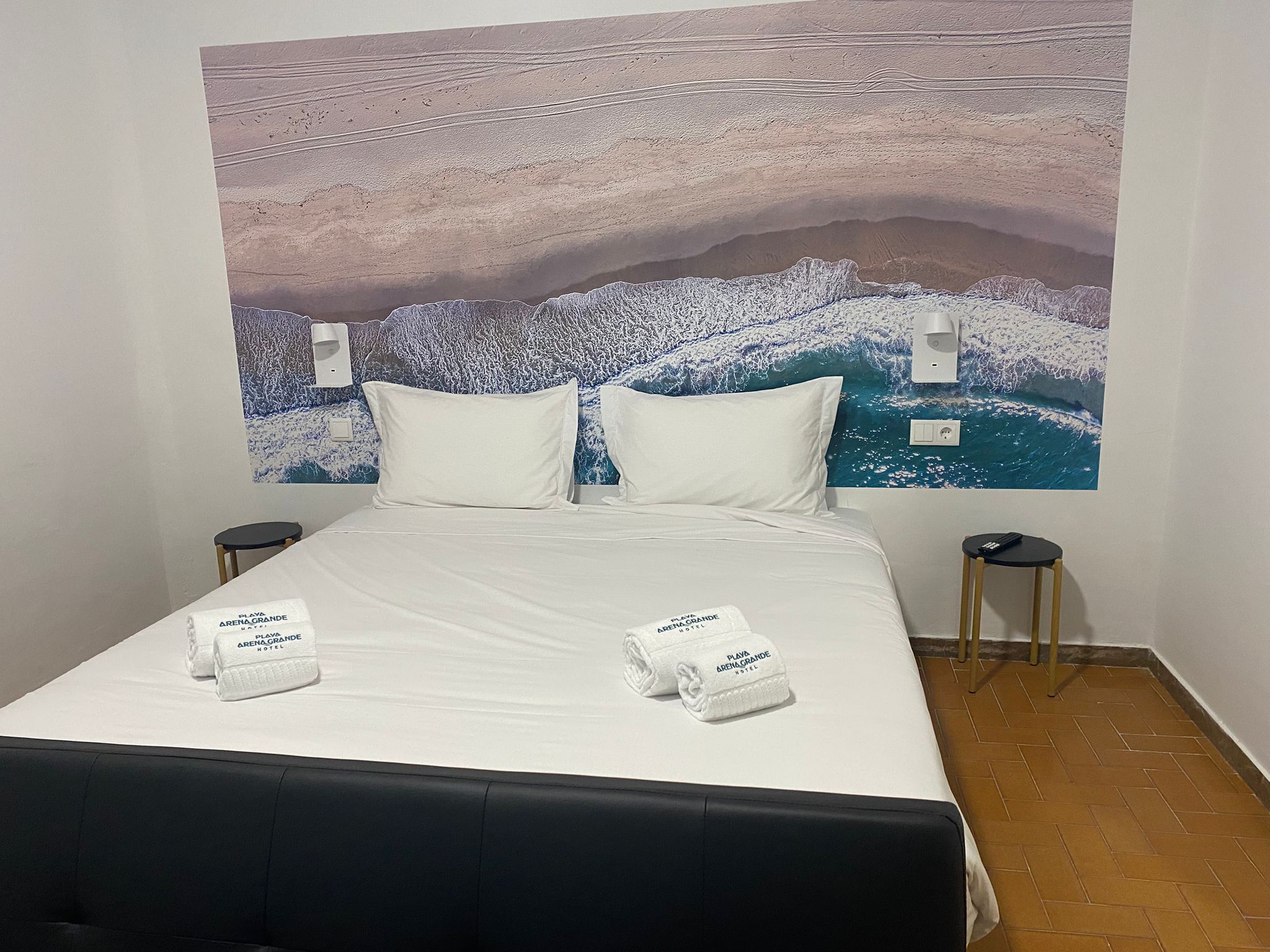 Quarto de hotel com cama, duas almofadas brancas, duas toalhas brancas, uma luminária na parede e uma mesinha com um controle remoto.