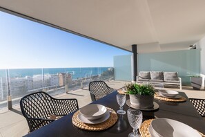 Uma área de refeições com mesa posta para quatro pessoas, com vista para a paisagem urbana e o mar.