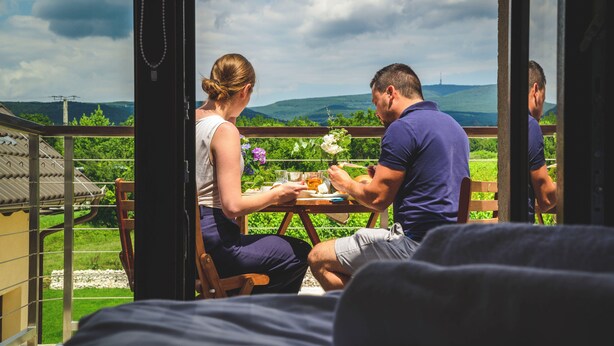 Mesa de jantar com duas pessoas sentadas, vista para colinas verdes e uma cama parcialmente visível.