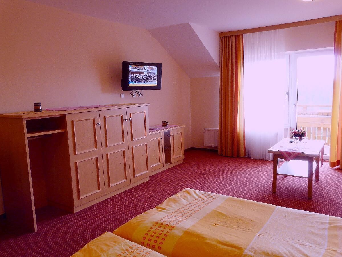 Quarto de hotel com cama, televisão na parede, armário de madeira, mesa pequena e varanda com cortinas.