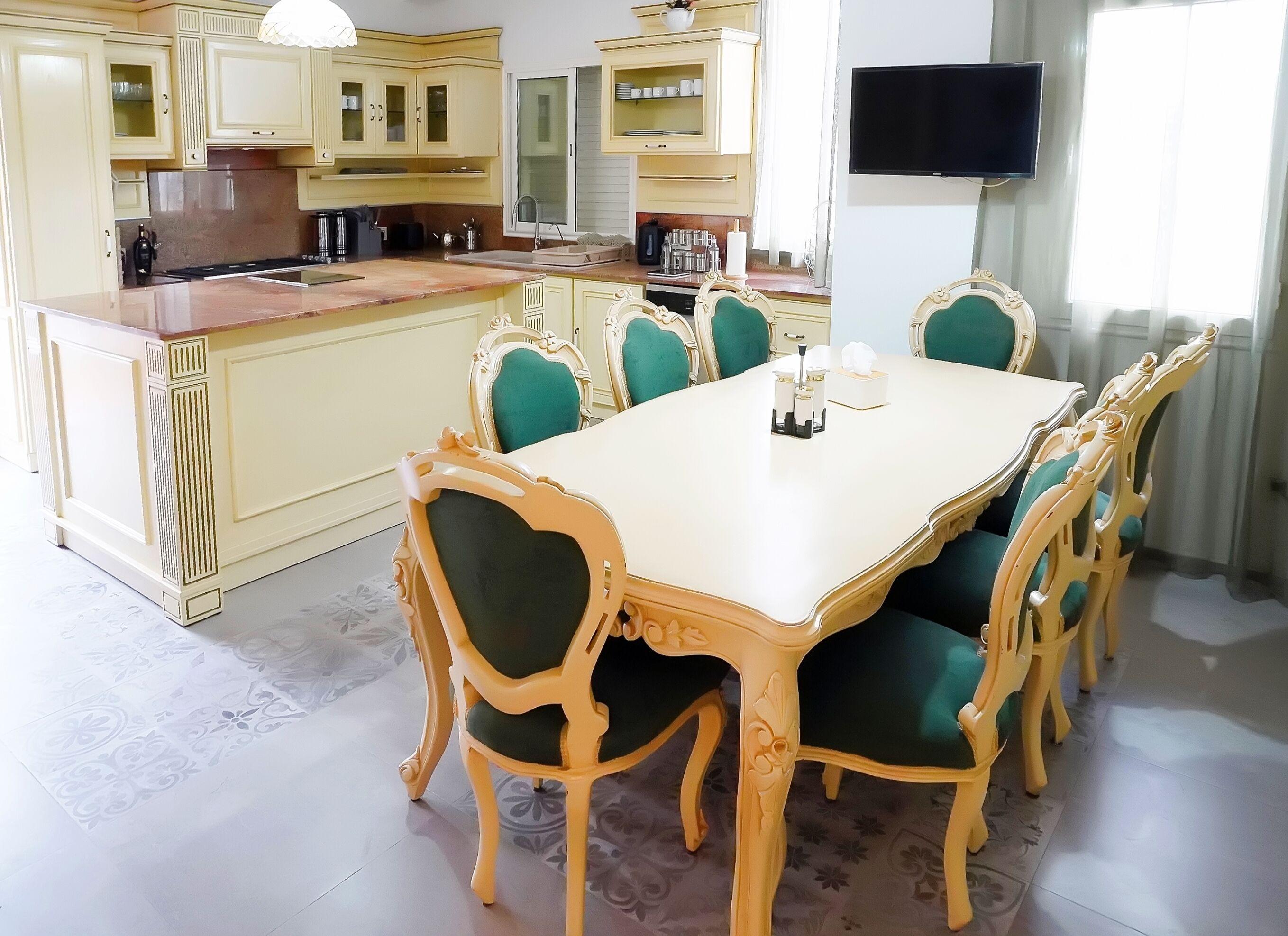 Uma sala de jantar com uma mesa grande e cadeiras, uma cozinha com armários amarelos e uma TV fixada na parede.