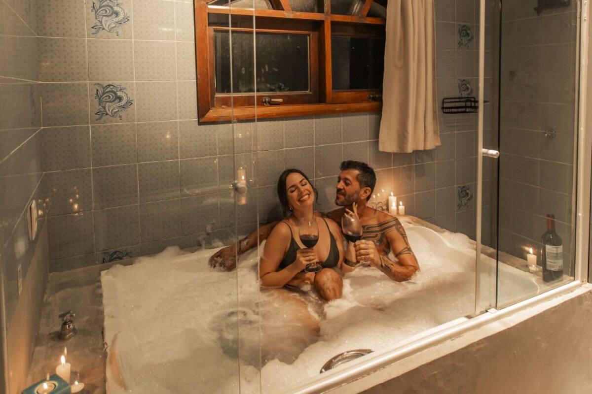Um casal em uma banheira de vidro, com velas, uma garrafa de vinho e uma janela acima.