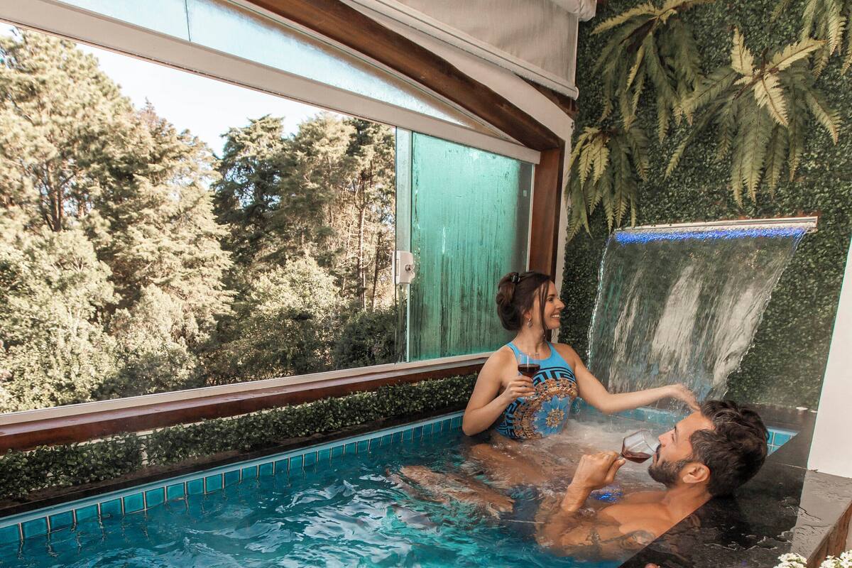 Área privativa com hidromassagem, cachoeira ao fundo, piscina e mesa com uvas.