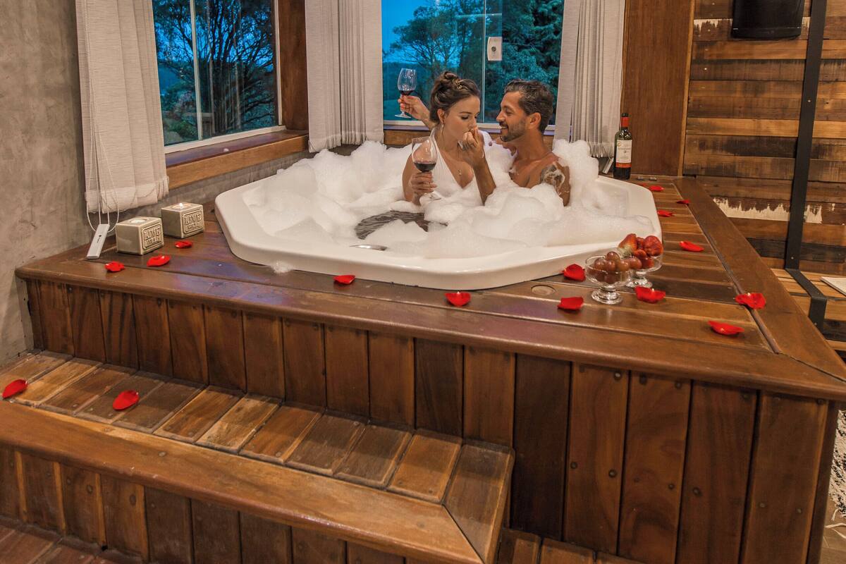 Um casal em uma banheira de hidromassagem com pétalas de rosas, vigas de madeira e vista para as árvores.