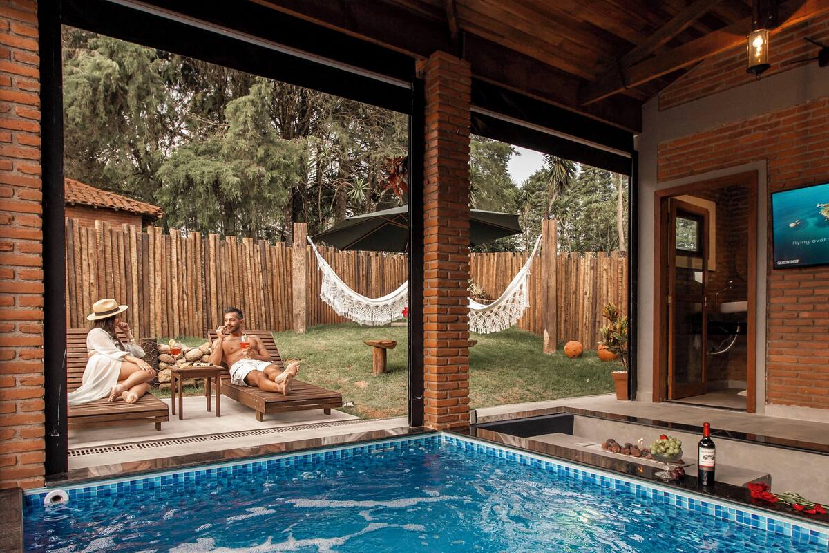 Piscina interna com teto de madeira, parede de tijolos e vista para um quintal com uma rede.