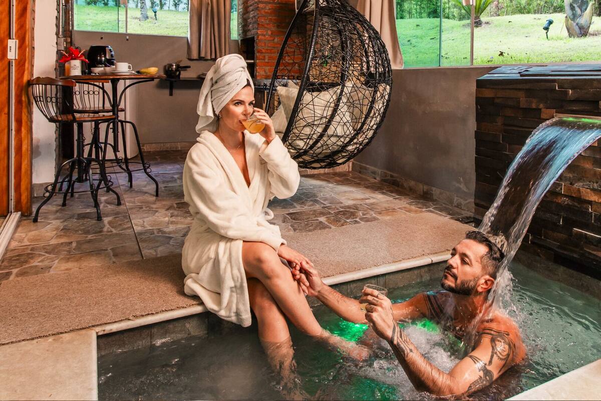 Um casal em uma banheira de hidromassagem com cachoeira, cercado por paredes de pedra e uma cadeira de vime.