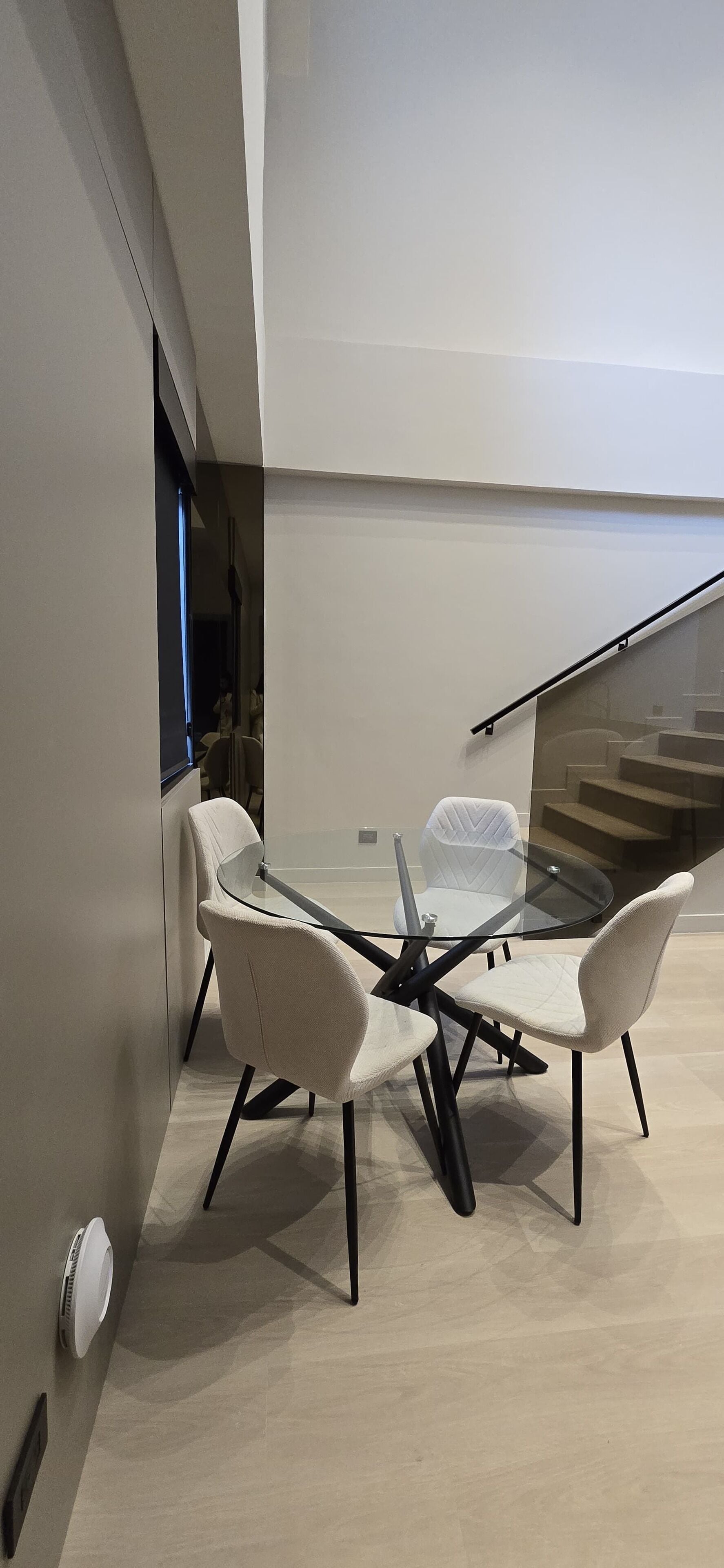 Uma sala de jantar moderna com mesa de vidro e quatro cadeiras brancas, uma escada ao fundo e um termostato fixado na parede.