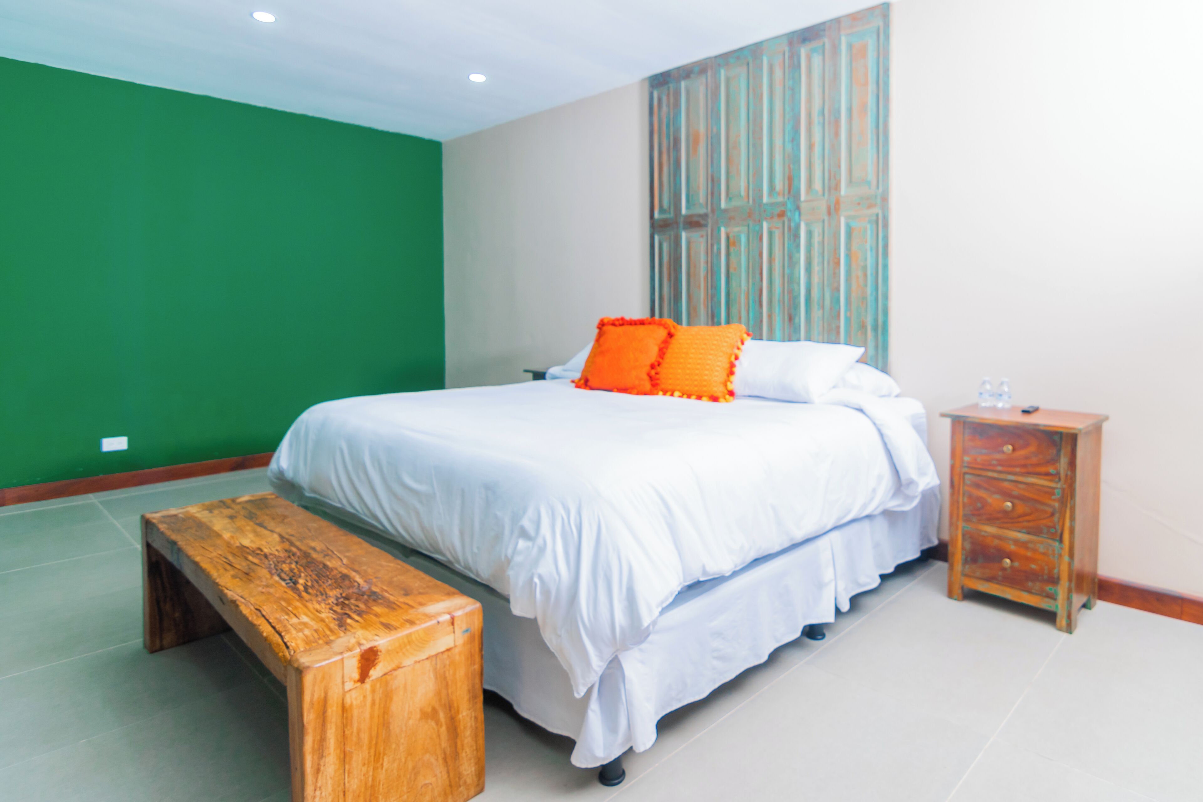 Quarto com cama, um banquinho de madeira, mesa de cabeceira e parede verde.