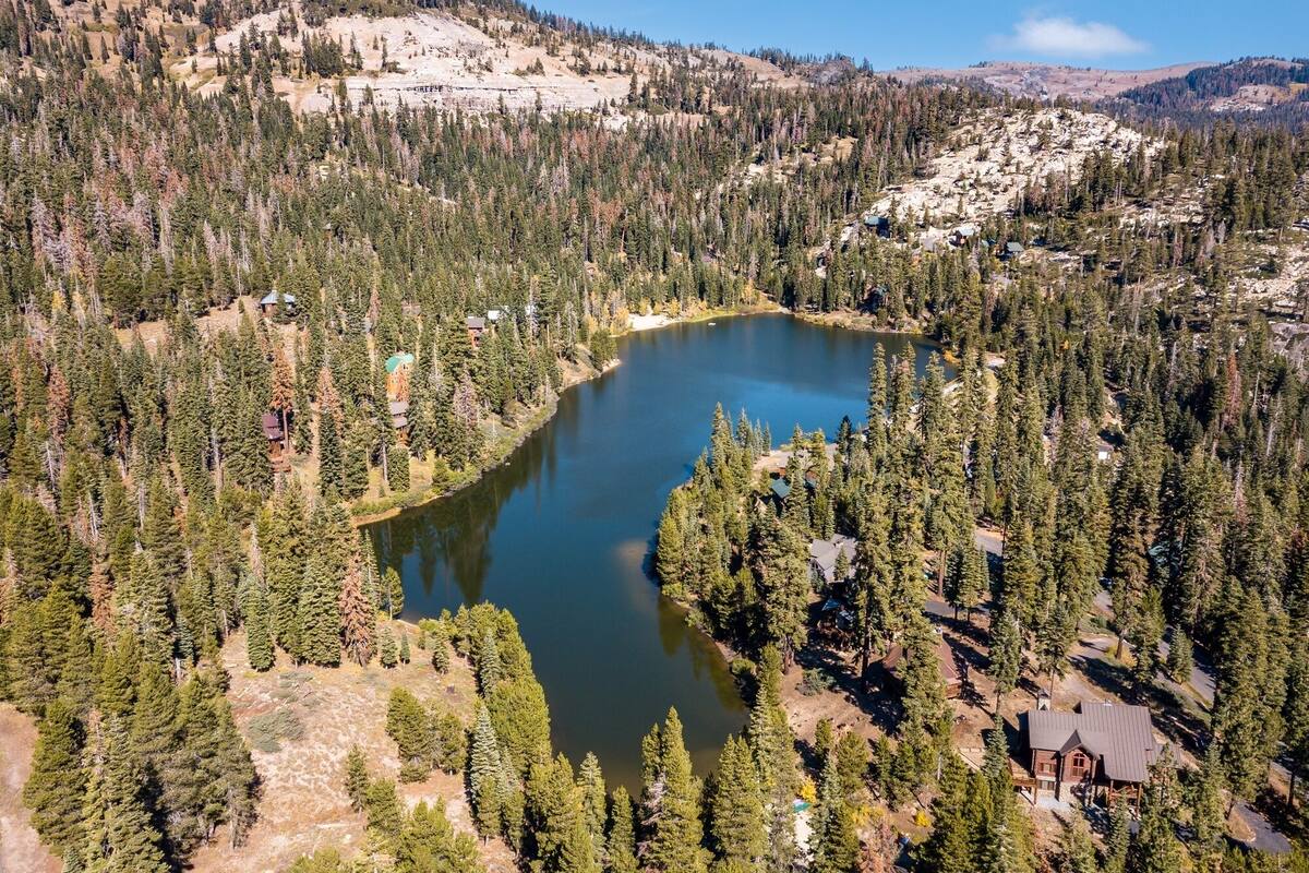 Uma pousada rodeada por floresta densa e um lago, com montanhas ao fundo.