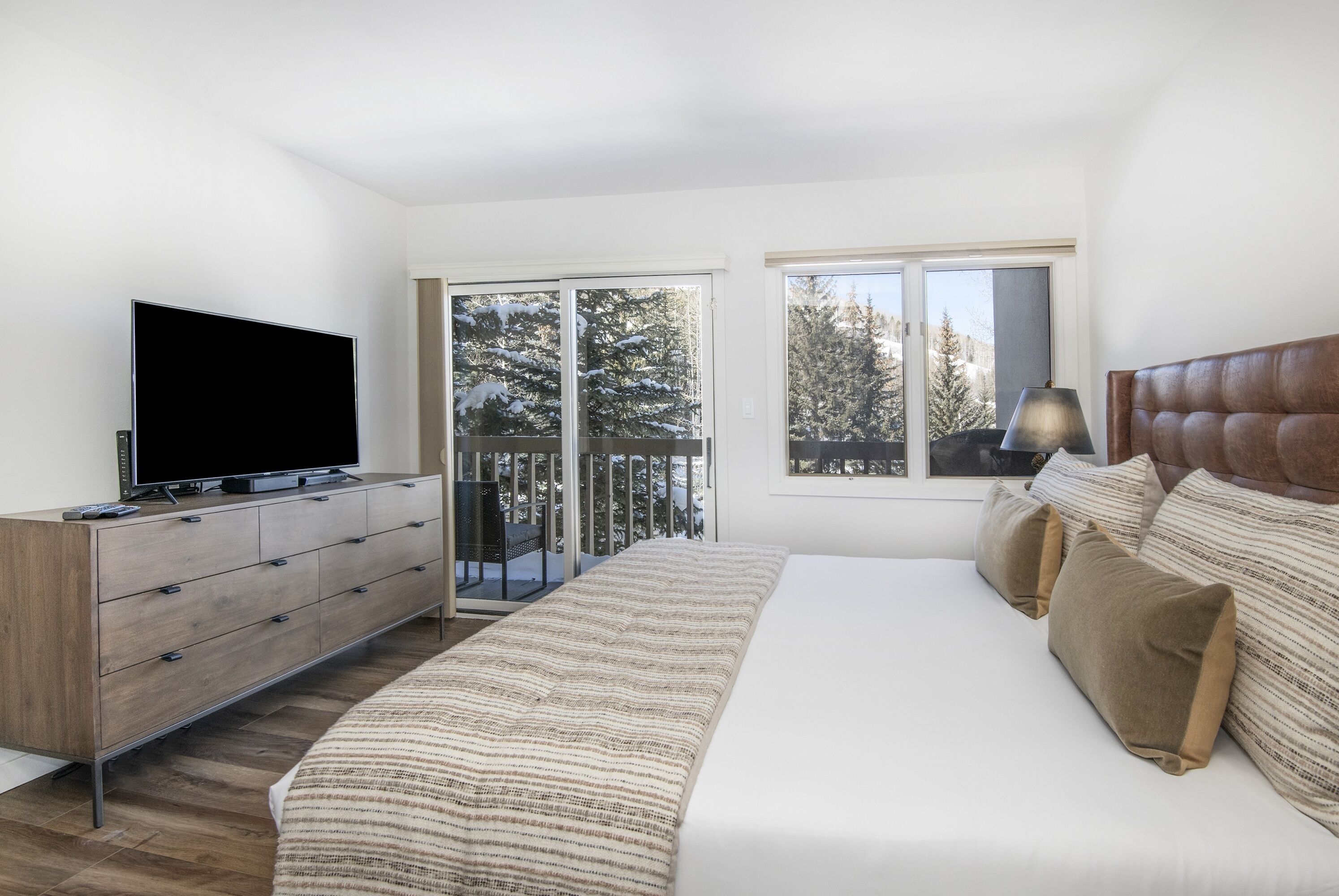 Quarto com cama grande, televisão em cômoda de madeira, varanda com vista para árvores e janela com vista para o exterior.