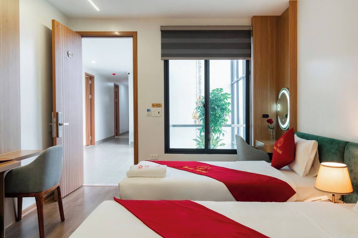 Quarto de hotel com duas camas, uma mesa, uma cadeira, janela com vista para o verde e um espelho na parede.