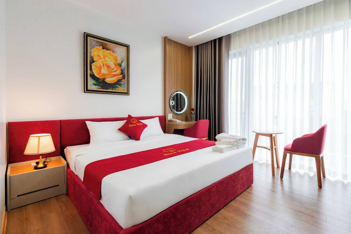 Quarto de hotel com uma cama grande, uma mesinha de cabeceira com abajur, uma cadeira vermelha, uma pequena mesa redonda e um quadro de uma rosa na parede.