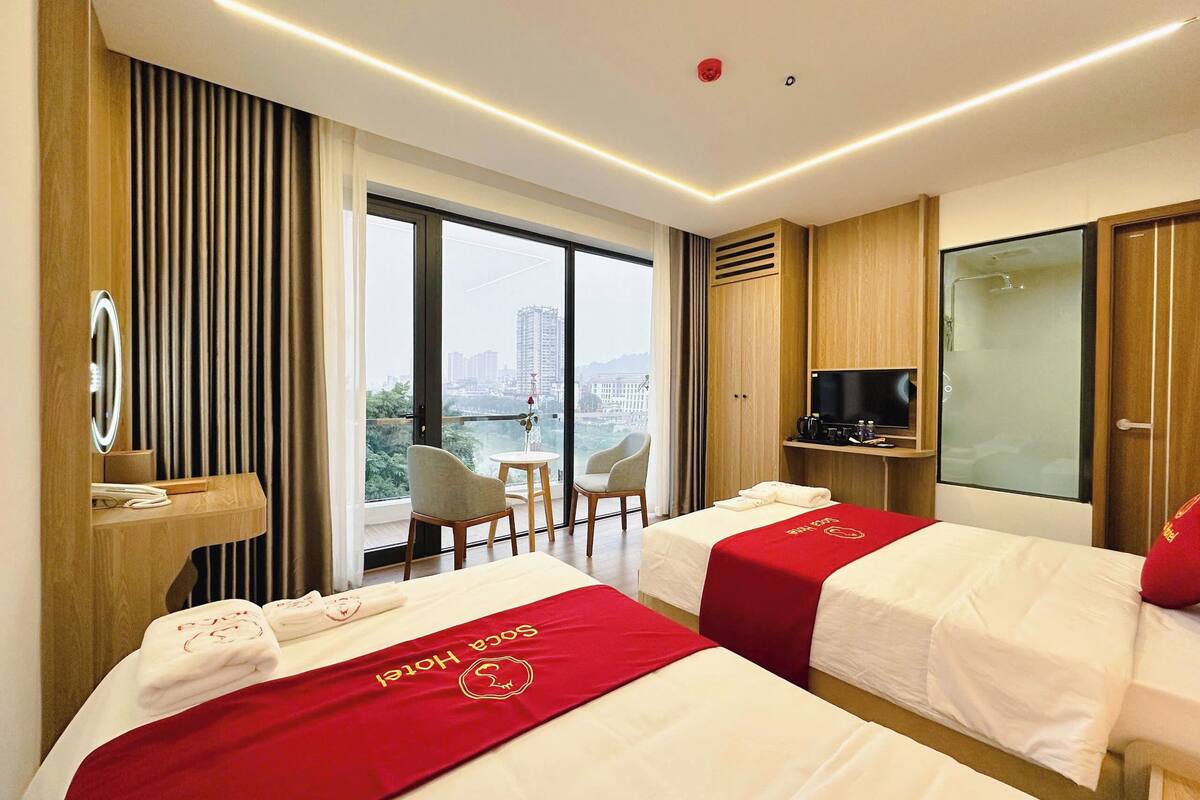 Quarto de hotel com duas camas, televisão, secretária e vista para a cidade.