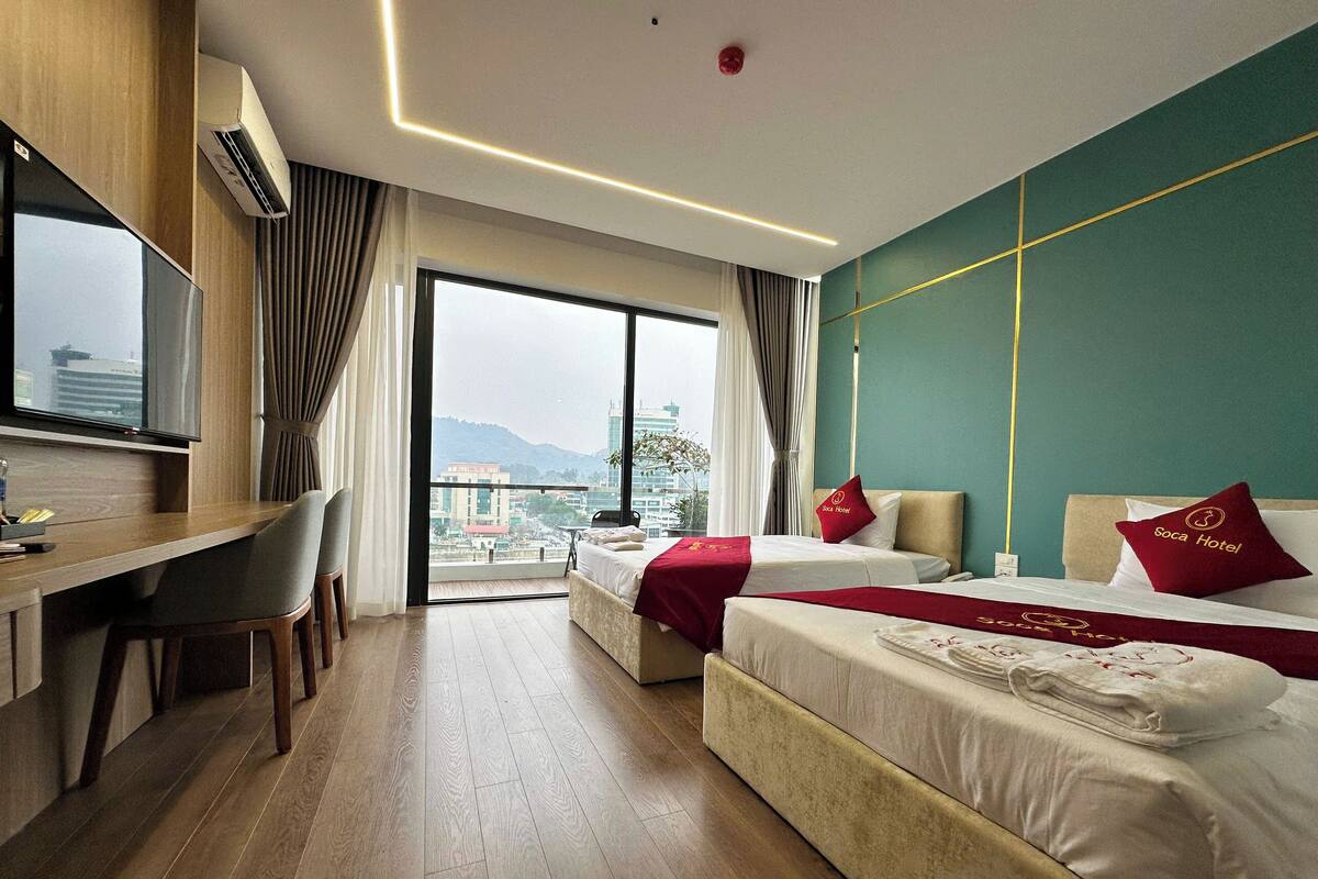Quarto de hotel com escrivaninha, duas camas, uma janela grande e vista para a paisagem urbana.