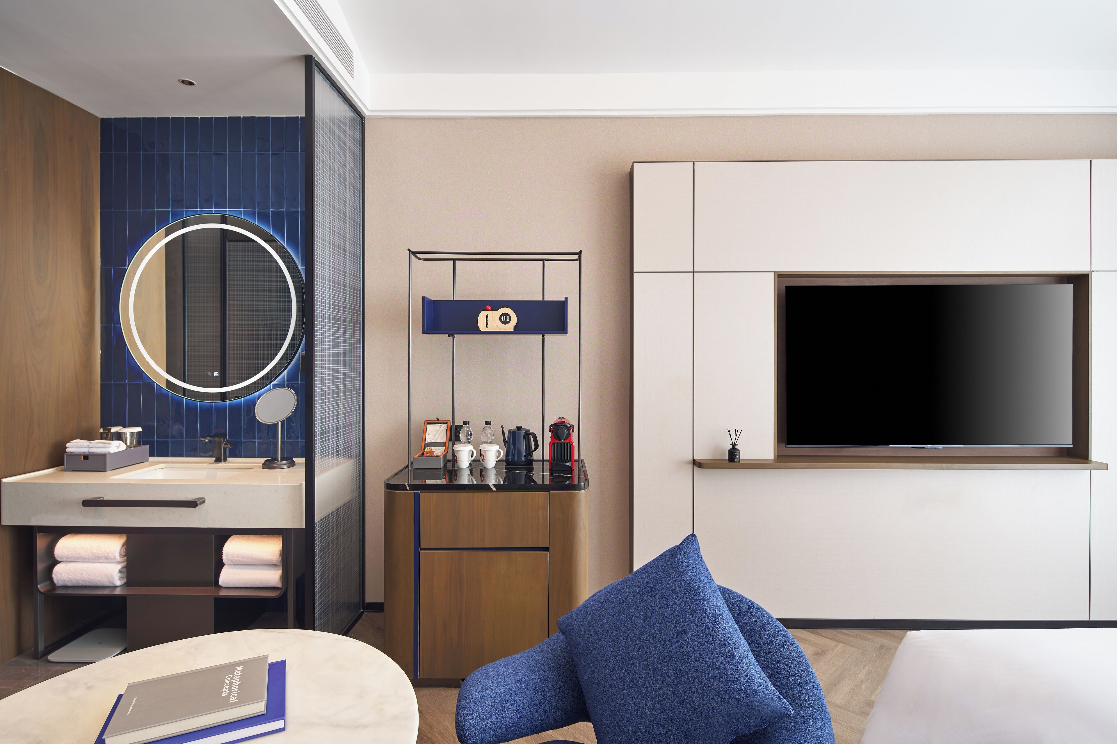 Quarto de hotel moderno com espelho redondo, TV de tela plana, armário de madeira e área de estar com almofada azul.
