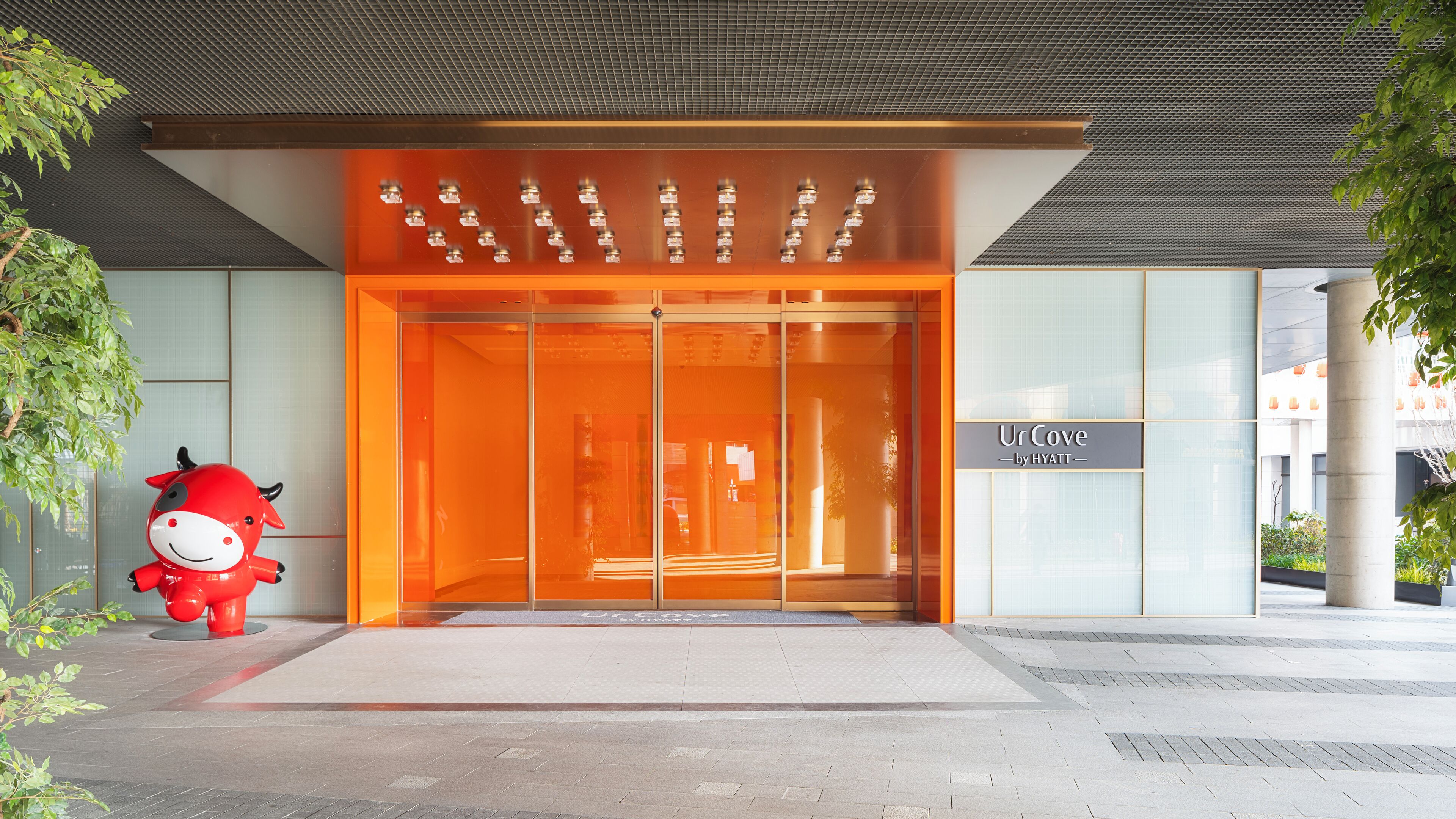 Entrada de um hotel com fachada moderna em laranja, porta de vidro e uma placa com a inscrição "Ur Cove by HYATT".