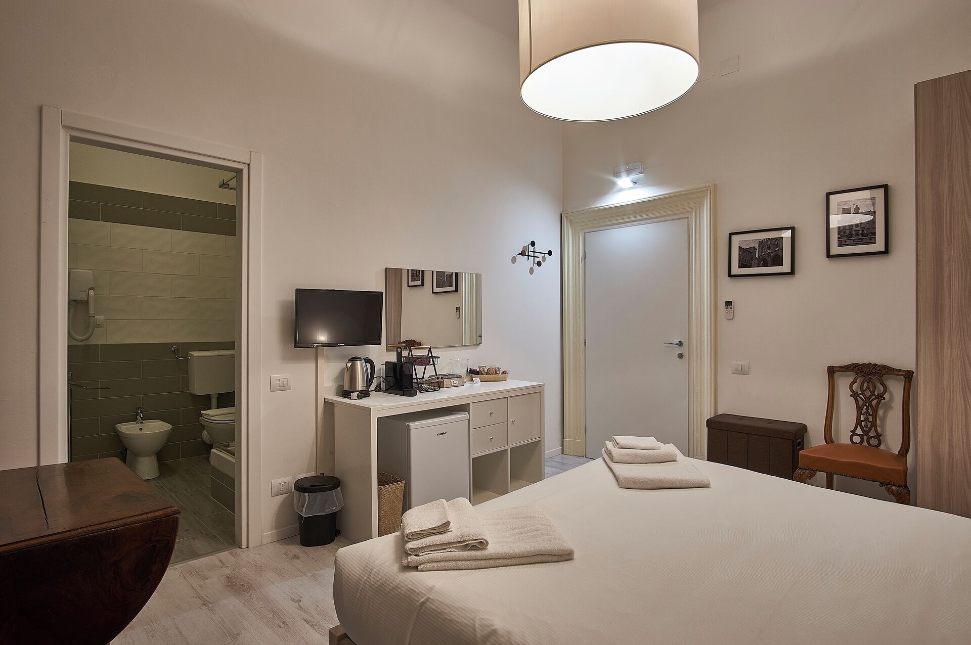 Quarto de hotel com cama, escrivaninha com televisão de tela plana, pequeno armário, lixeira e porta que dá para outro quarto.