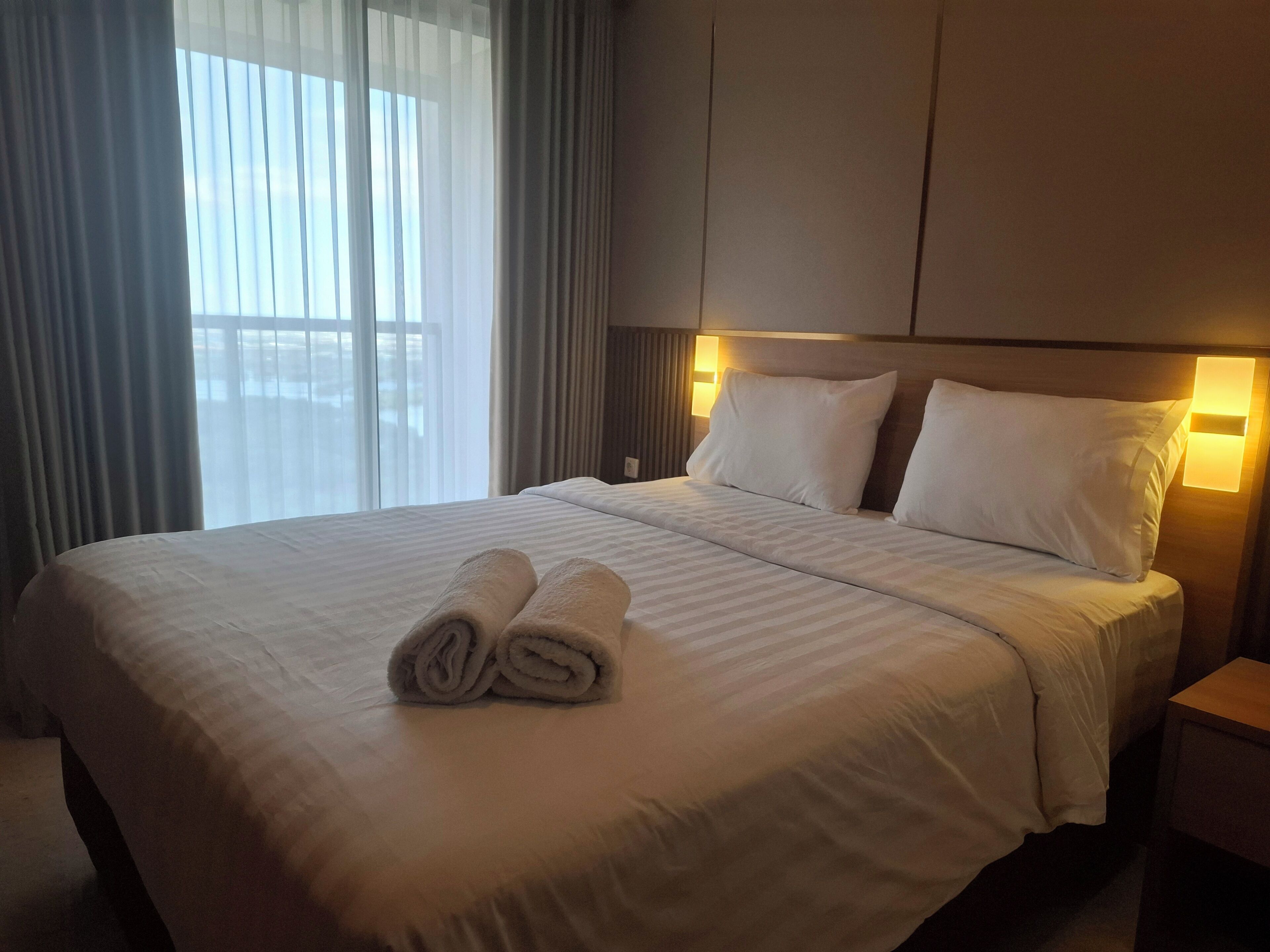 Quarto de hotel com cama bem arrumada, duas almofadas, cabeceira com iluminação integrada e janela com cortinas.