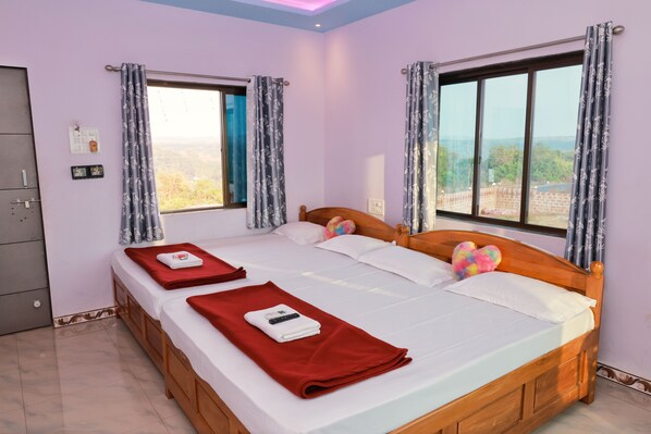 Uma cama de casal com roupa de cama vermelha e branca, duas janelas com cortinas estampadas e vista para a paisagem.