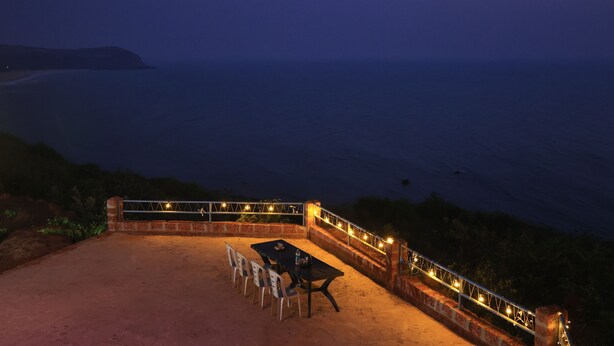 Área de refeições ao ar livre com mesa e cadeiras, com vista para uma praia e oceano à noite.