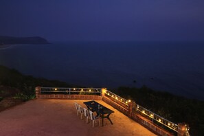 Área de refeições ao ar livre com mesa e cadeiras, com vista para uma praia e oceano à noite.