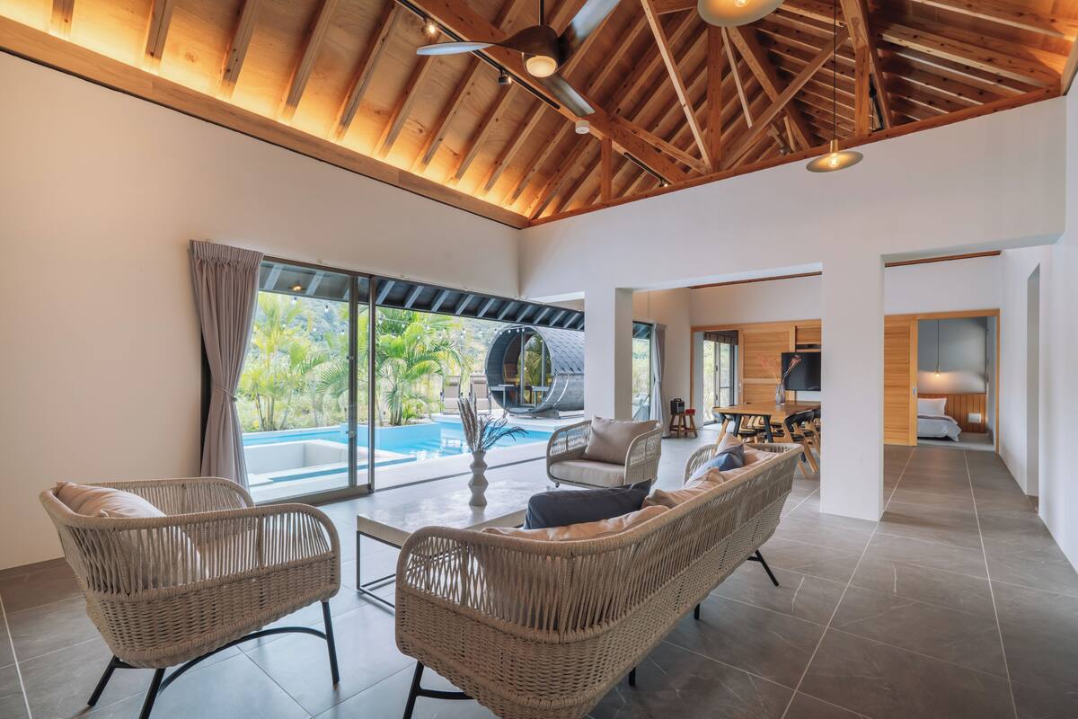 Ampla sala de estar com móveis de vime, vista para piscina e cozinha moderna ao fundo.