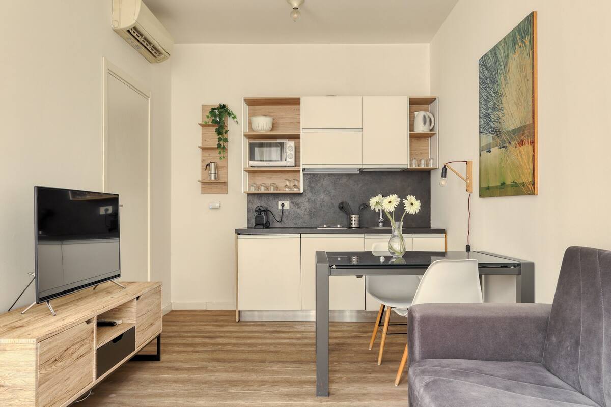 Uma sala de estar moderna com televisão de tela plana, um sofá cinza, uma mesa de jantar com cadeiras e uma área de cozinha com micro-ondas, chaleira e prateleiras.