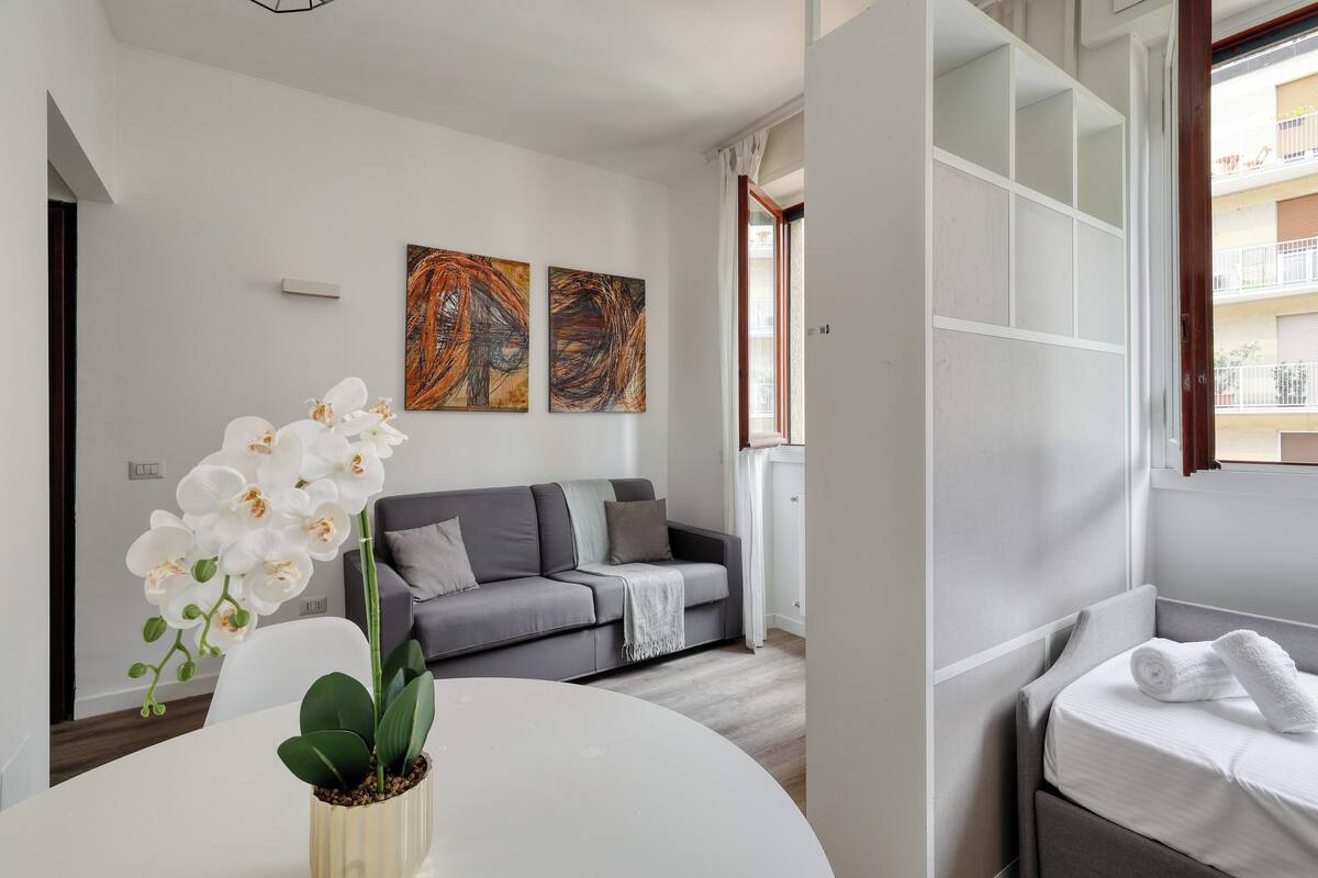 Uma sala de estar moderna com um sofá cinzento, uma mesa branca com uma planta orquídea e arte abstrata na parede.