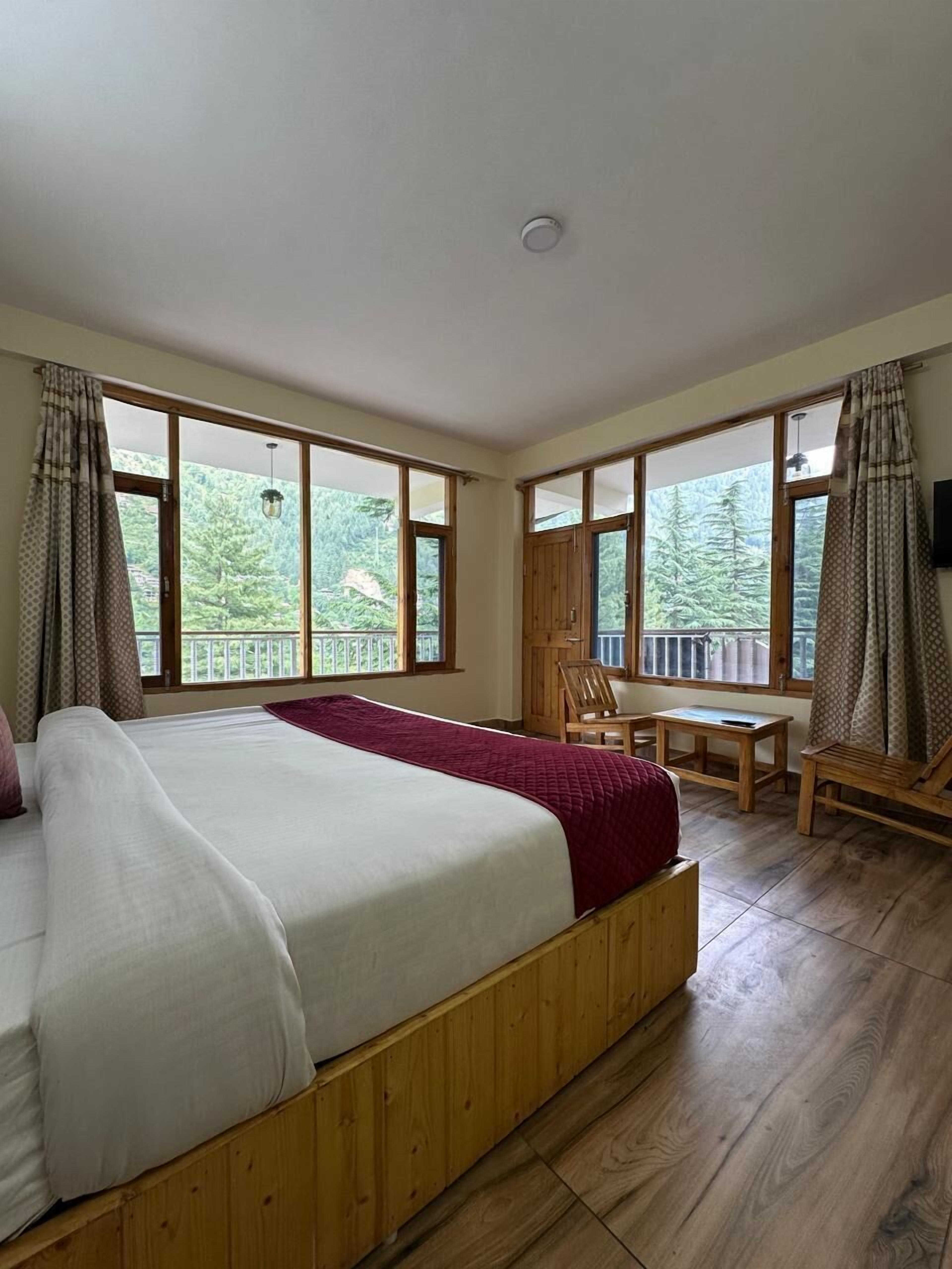 Quarto com uma cama grande, uma escrivaninha de madeira e uma cadeira. Há janelas amplas com cortinas.