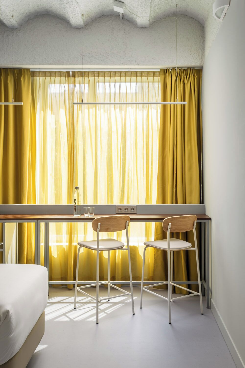 Quarto com cama, escrivaninha e duas cadeiras, além de janela com cortinas amarelas.