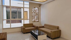 Sala de estar com sofá bege, poltrona combinando e mesa de centro. Há uma janela grande com um painel decorativo e luminária fixada na parede.
