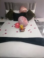 Cama com lençol branco, balões rosa e vermelhos, e uma pequena cesta de presentes.