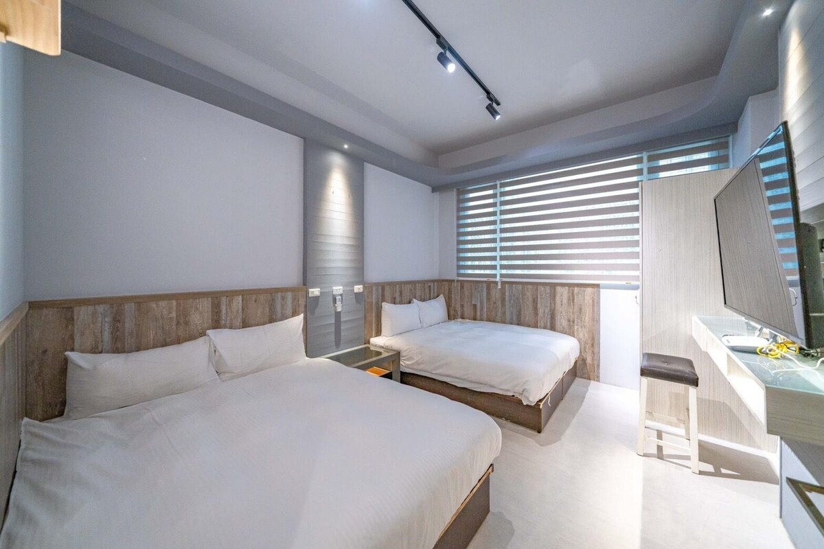 Quarto de hotel moderno com duas camas, uma escrivaninha e uma janela grande com persianas.