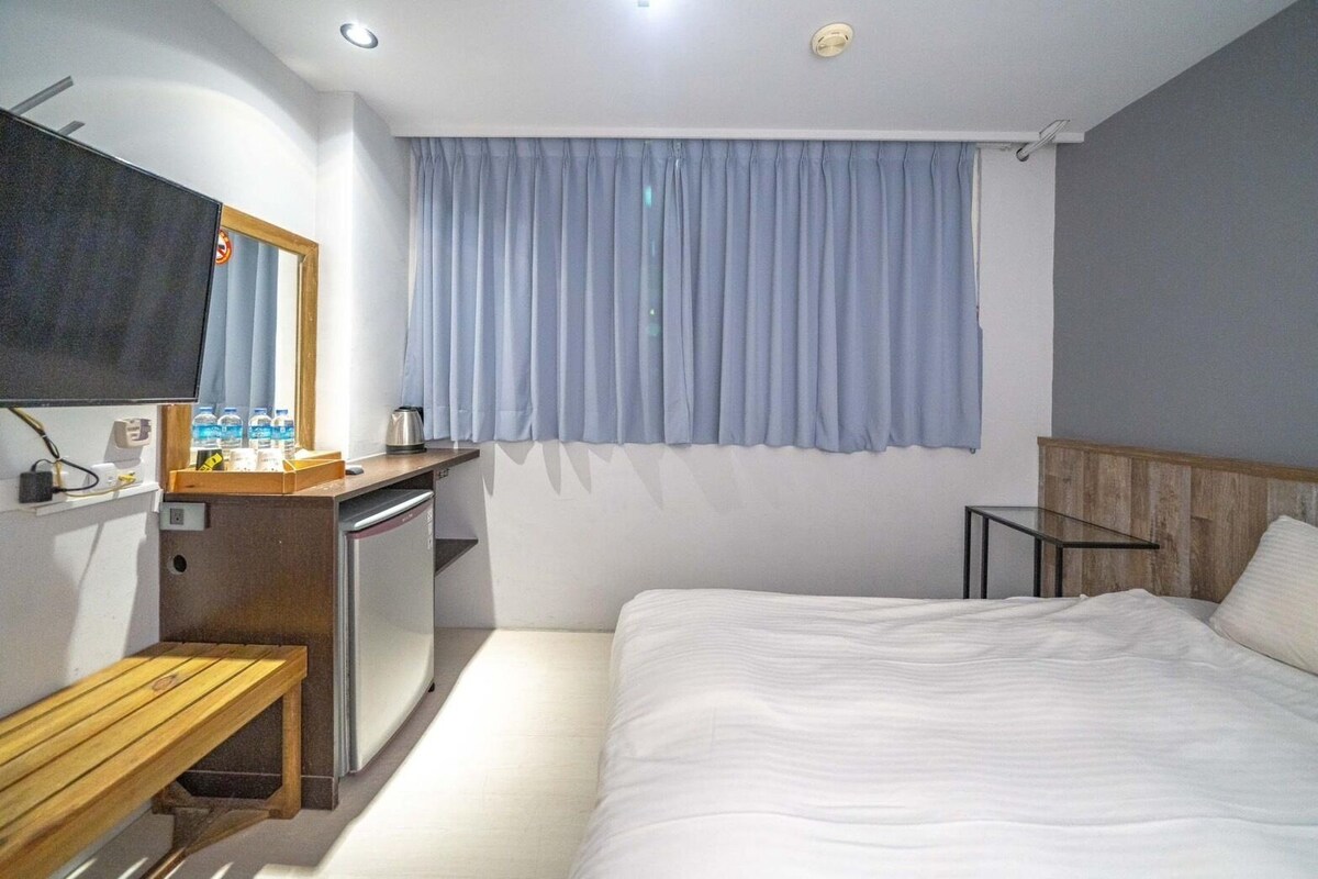 Quarto de hotel com cama, escrivaninha, espelho, pequeno espaço de cozinha e janela com cortinas.