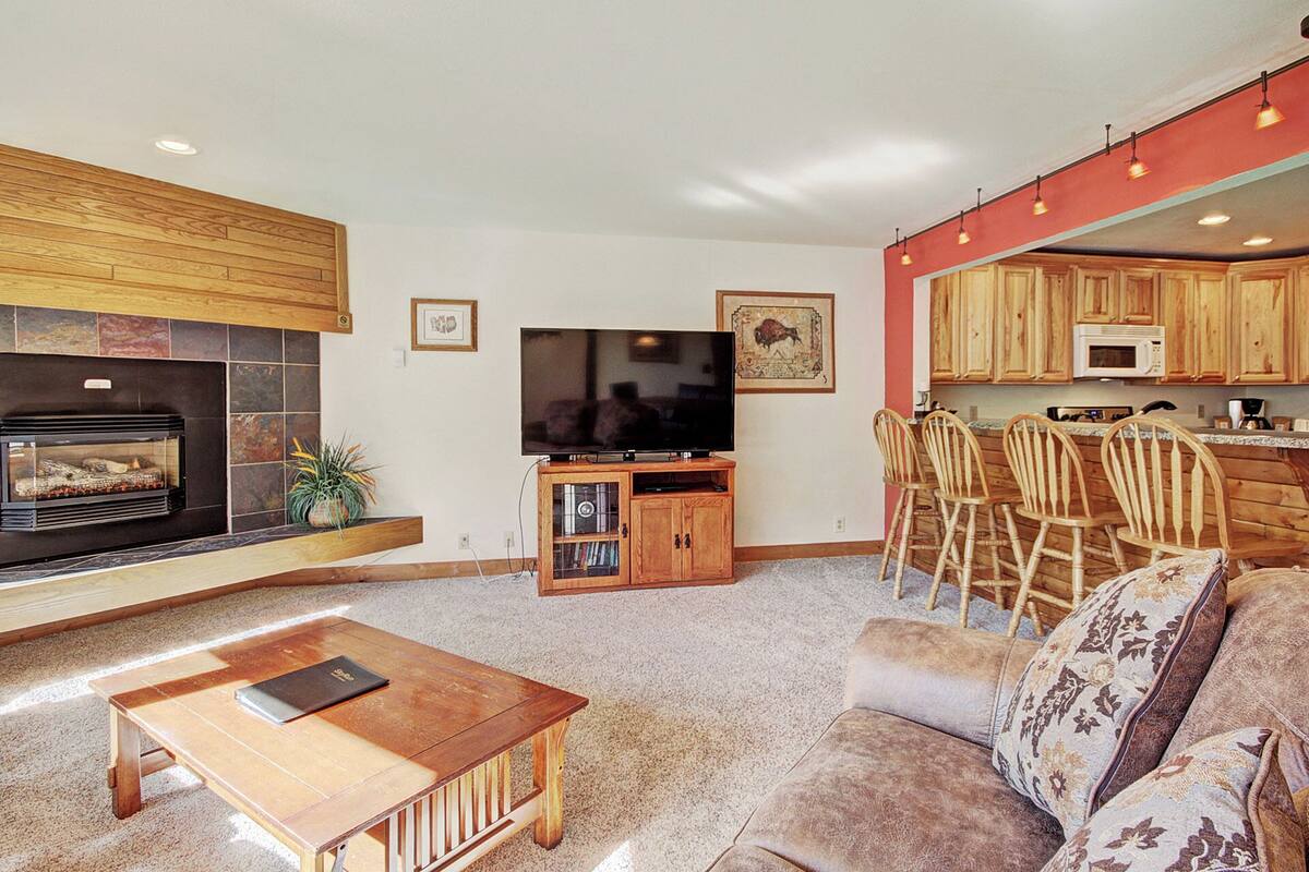 Sala de estar com lareira, TV e sofá. Há uma cozinha com armários de madeira e micro-ondas.