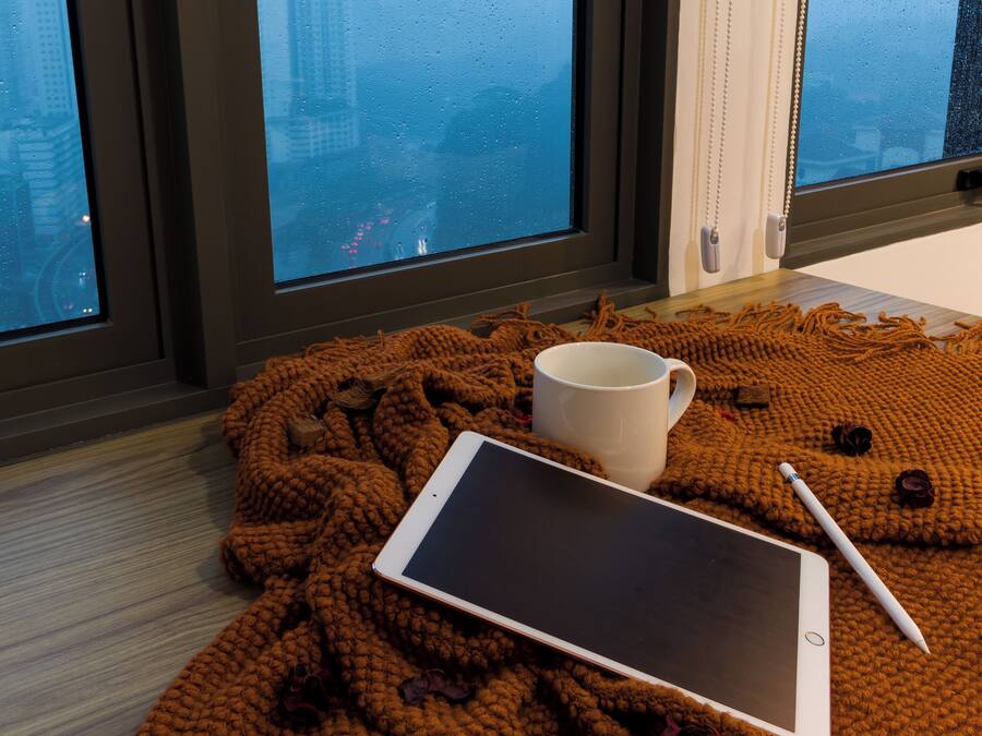 Ambiente acolhedor em interiores com um tablet, uma caneca e uma caneta sobre um cobertor de malha, com uma janela exibindo uma vista urbana.