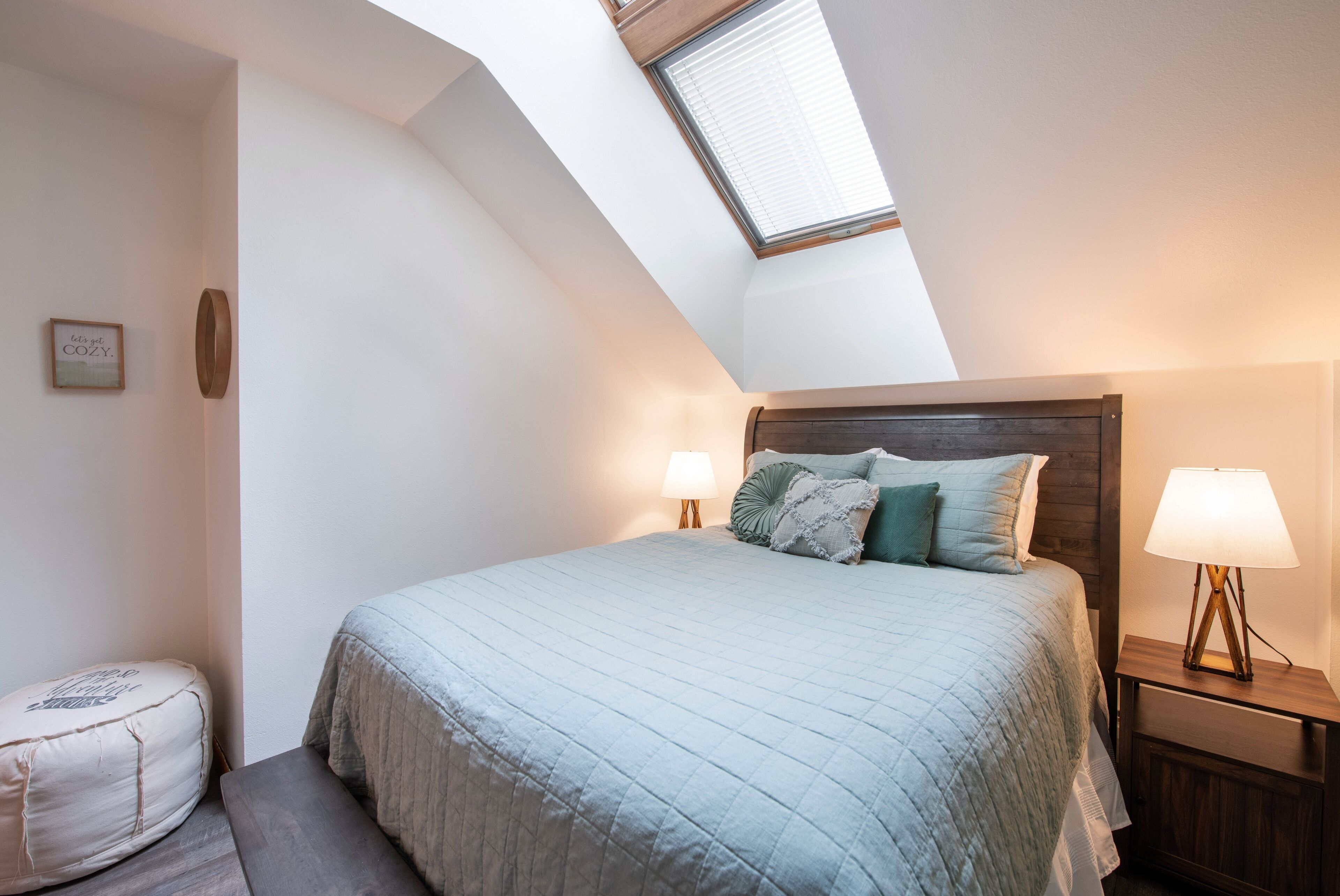 Um quarto com teto inclinado e claraboia, uma cama com colcha acolchoada, duas luminárias de cabeceira e um banquinho redondo pequeno.