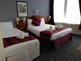Quarto de hotel com duas camas, cada uma com colcha listrada em vermelho e preto, almofadas e toalhas brancas. Há um quadro pendurado na parede e uma janela com cortinas.