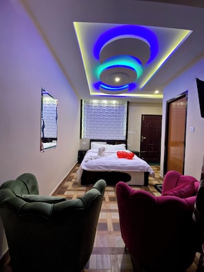 Quarto de hotel com cama, televisão fixada na parede, teto com iluminação circular e duas cadeiras.